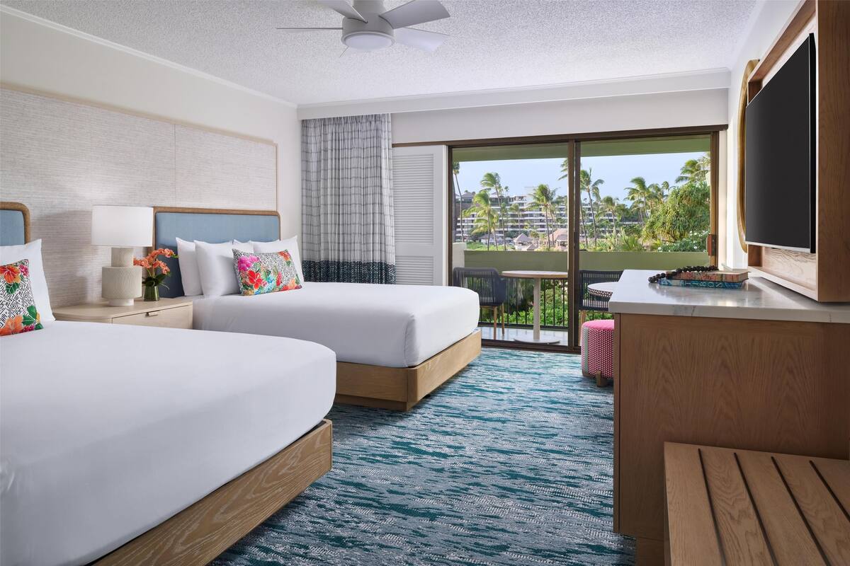Quarto de hotel com duas camas, uma escrivaninha, televisão, varanda com vista para palmeiras e um ventilador de teto.