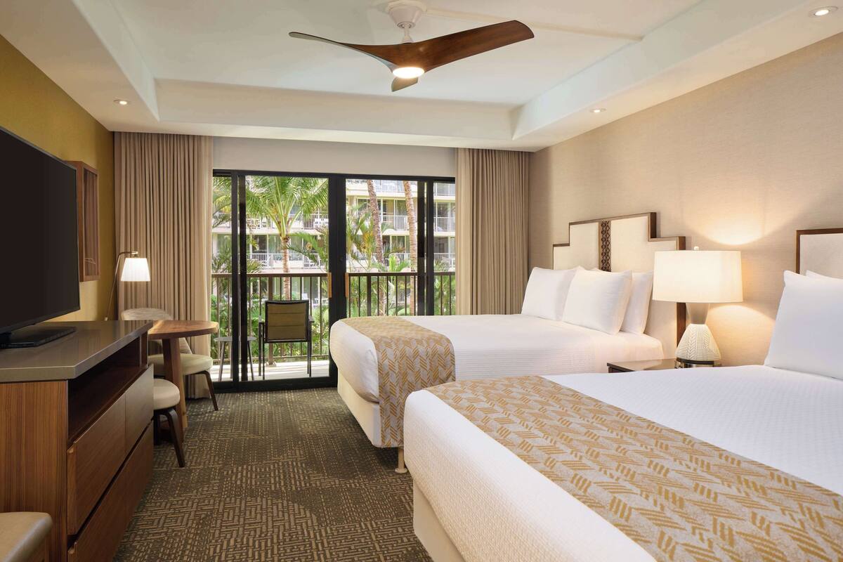 Quarto de hotel com duas camas, uma escrivaninha com televisão, um ventilador de teto e vista para palmeiras e edifícios.