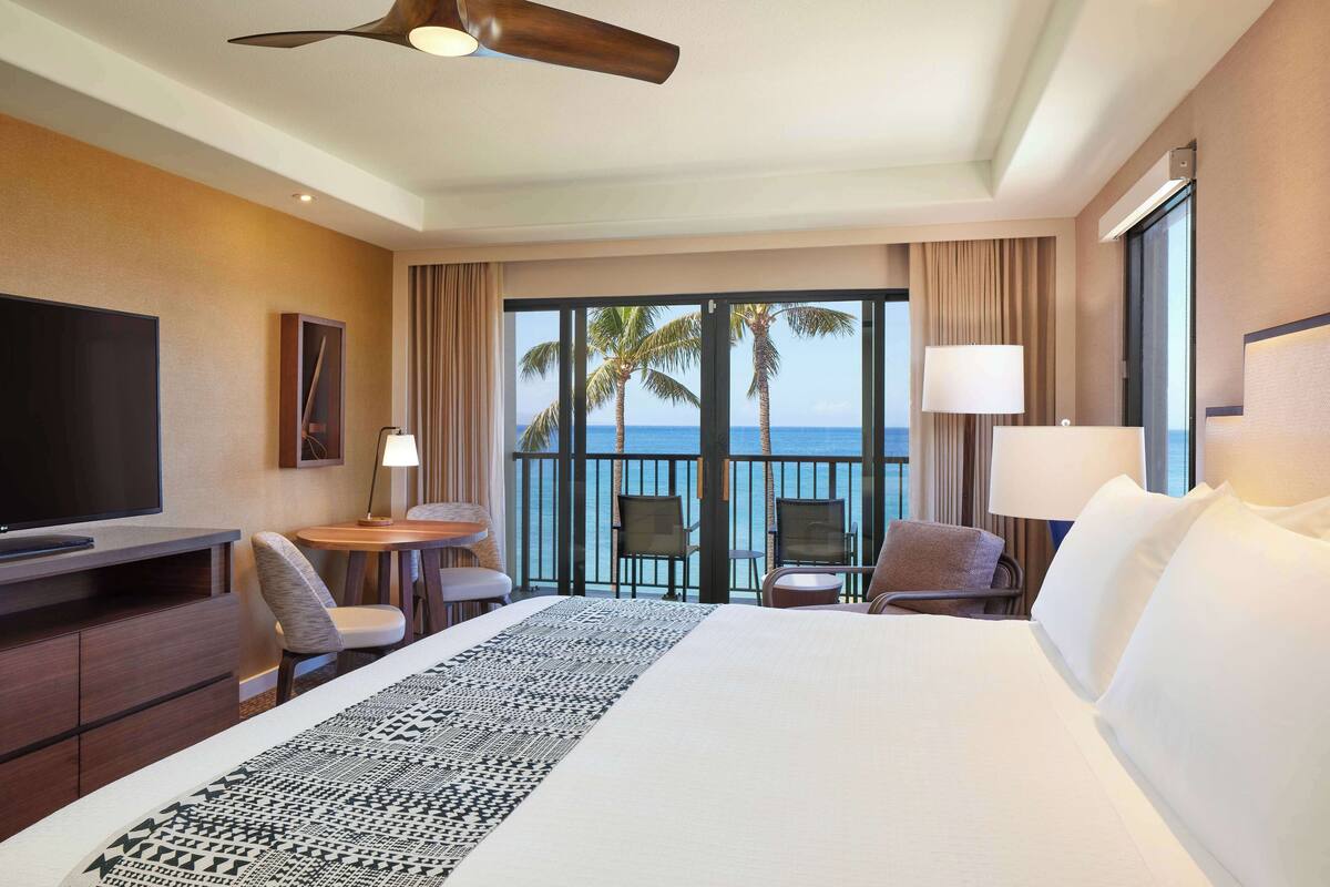 Quarto de hotel com cama, televisão, escrivaninha, cadeira, luminária e vista para o oceano.