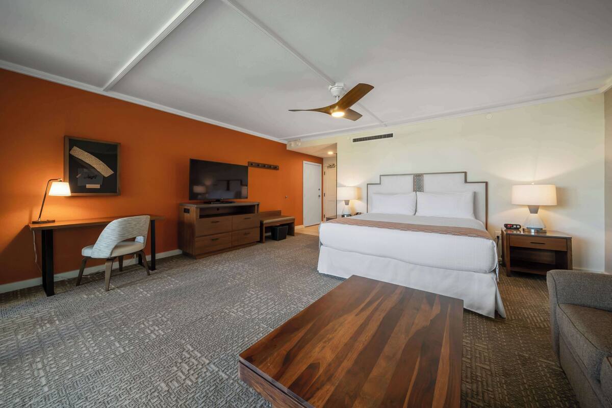 Quarto de hotel com cama, escrivaninha, televisão, ventoinha no teto e piso coberto por carpete.