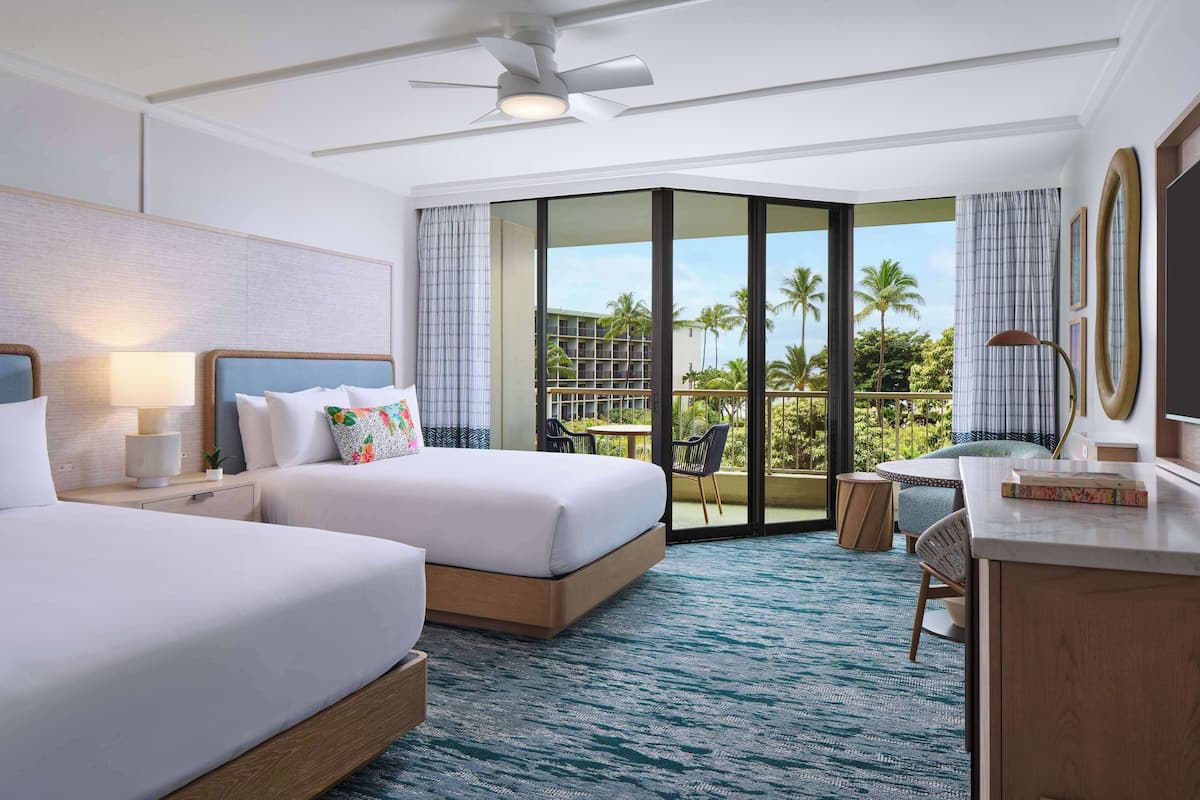 Quarto de hotel com duas camas, uma mesa, uma cadeira, varanda com vista para palmeiras e um ventilador de teto.