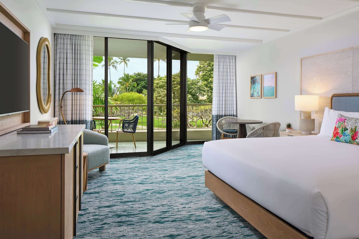 Quarto de hotel com cama, escrivaninha, cadeira, varanda e vista para vegetação, além de um ventilador de teto.