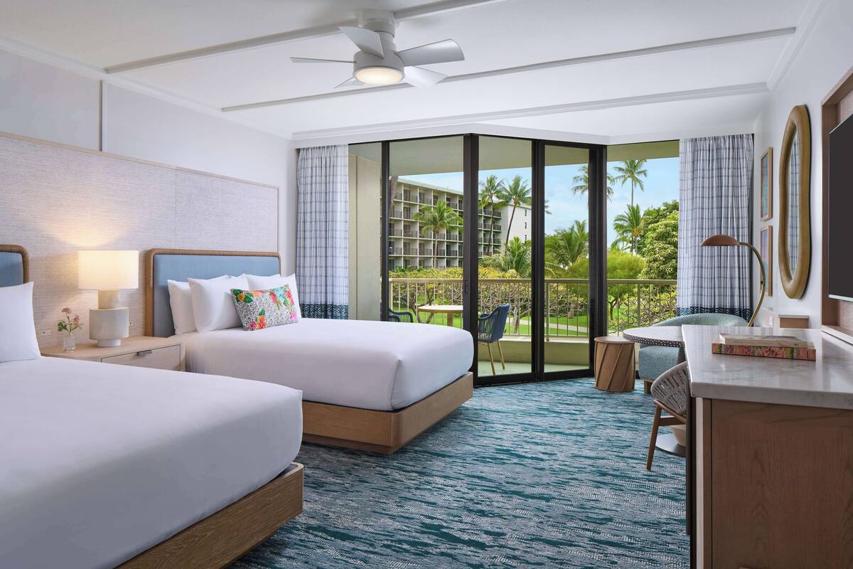 Quarto de hotel com duas camas, uma escrivaninha, uma cadeira, ventoinha no teto e vista para palmeiras e um edifício.