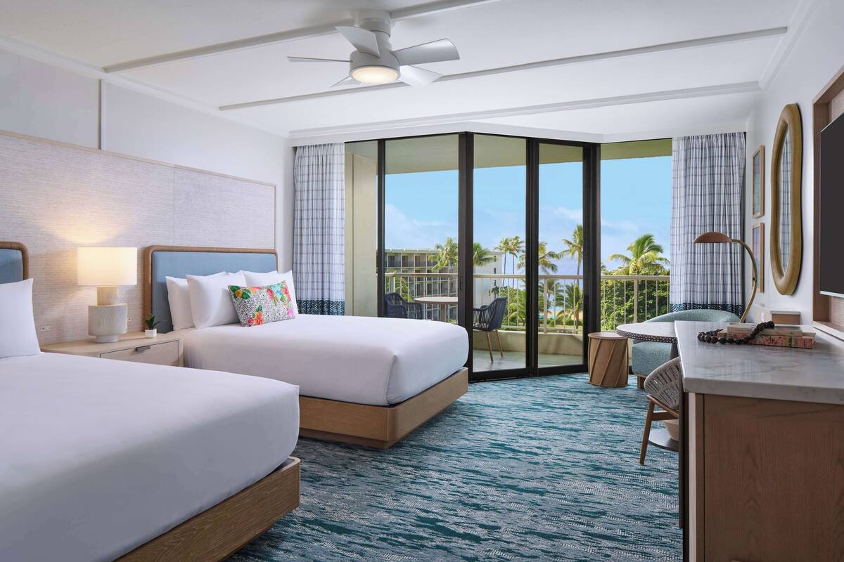 Quarto de hotel com duas camas, uma escrivaninha, uma cadeira, um ventilador de teto e uma varanda com vista para palmeiras.