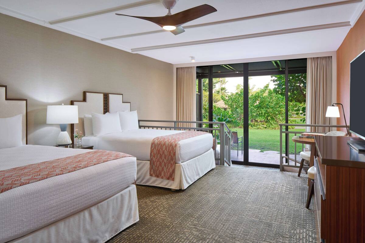 Quarto de hotel com duas camas, uma escrivaninha, televisão e varanda com vista para vegetação.