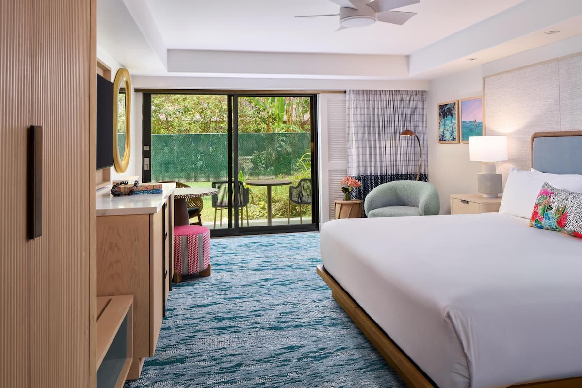 Quarto de hotel com cama, escrivaninha, cadeira, mesinha, luminária e vista para o exterior.