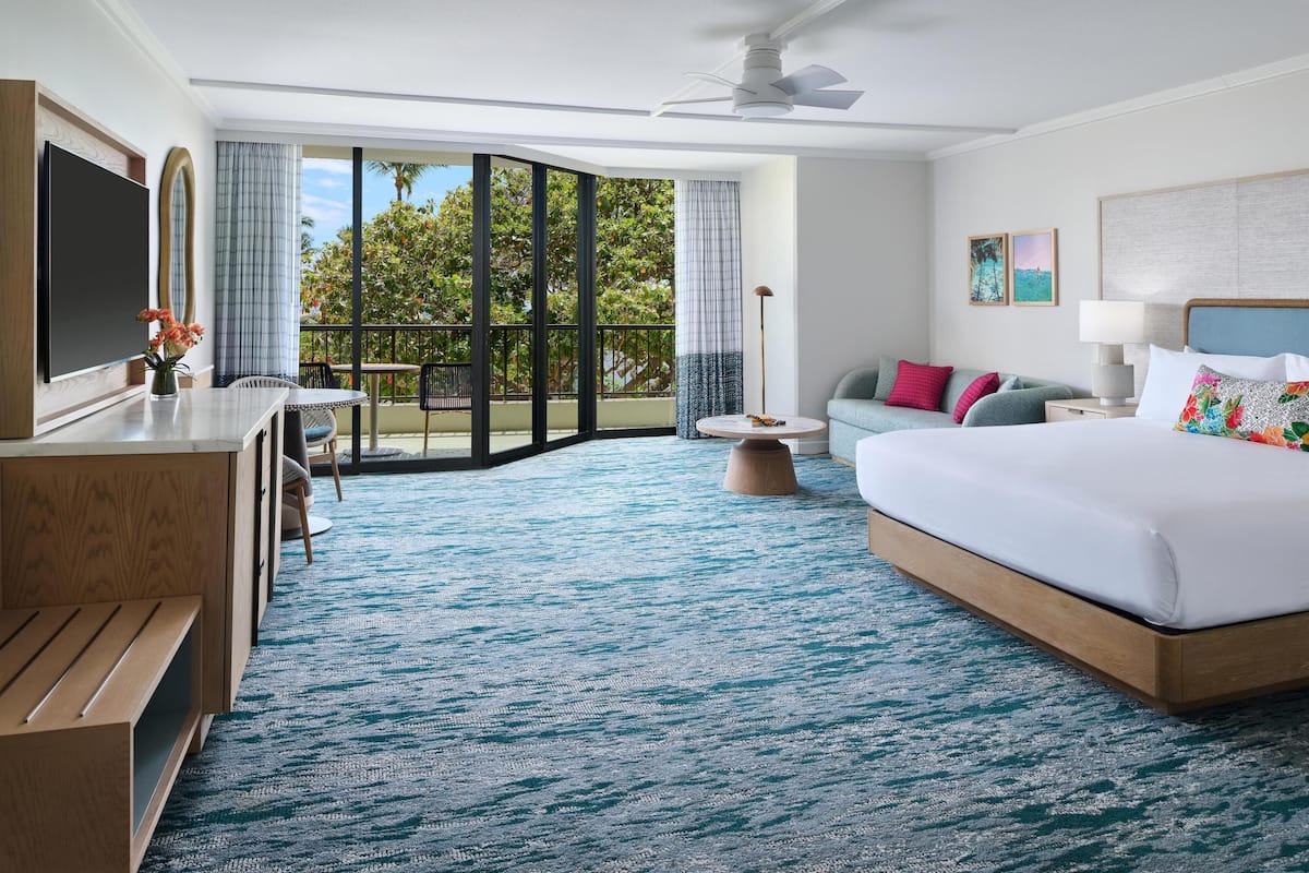 Quarto de hotel com uma cama grande, escrivaninha, cadeira, sofá, mesa redonda pequena, televisão e varanda com vista para árvores.