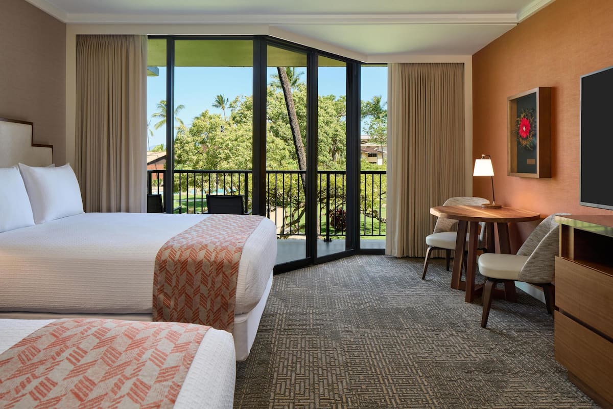 Quarto de hotel com uma cama grande, uma escrivaninha, uma cadeira e vista para palmeiras através de uma varanda.