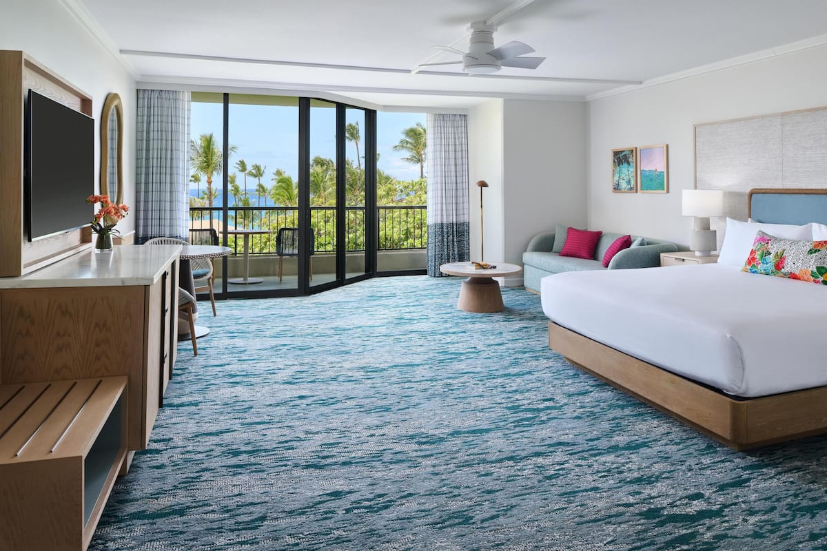Quarto de hotel com uma cama grande, uma mesa de escritório, uma cadeira, uma mesinha e uma varanda com vista para palmeiras.