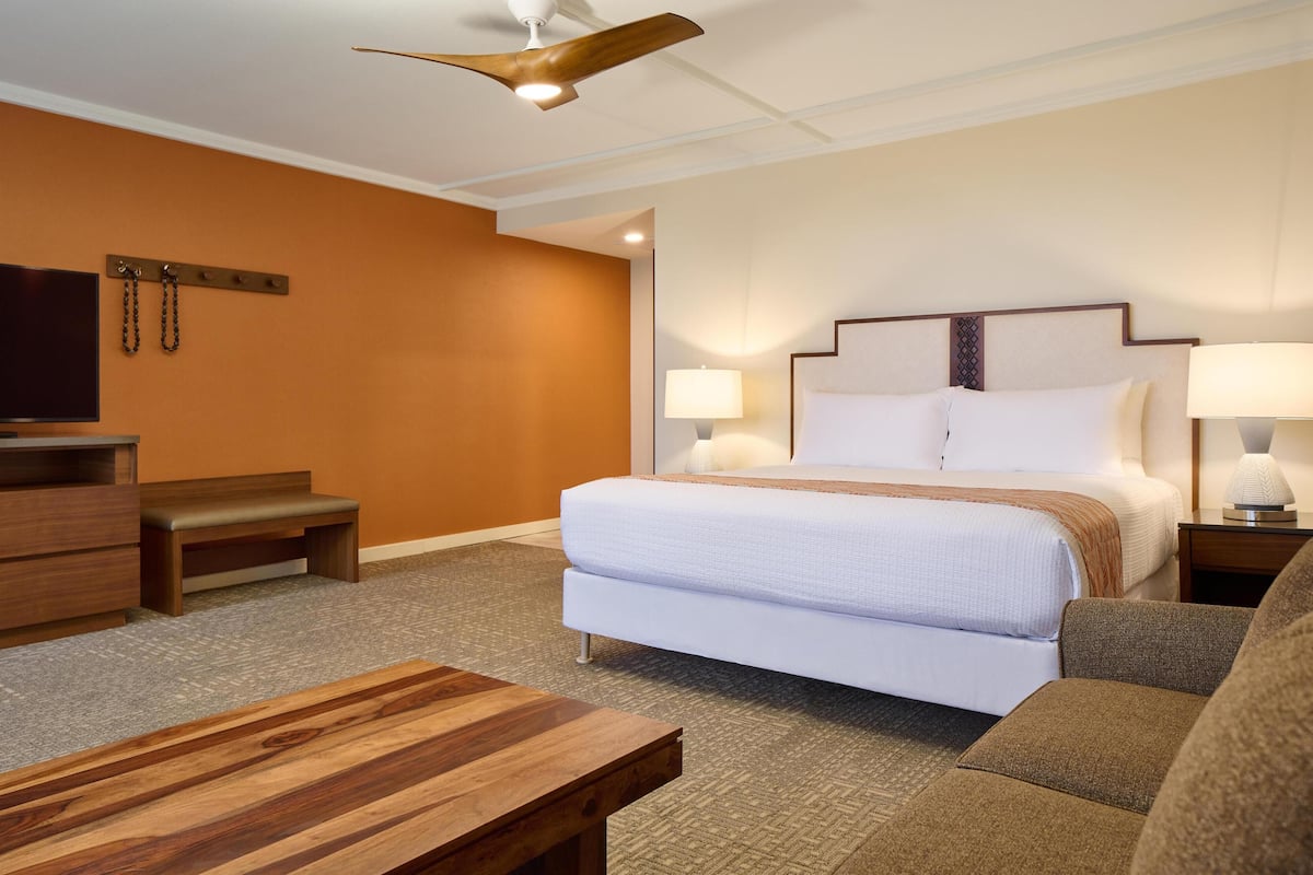 Quarto de hotel com cama, abajures, um banco de madeira, escrivaninha e ventilador de teto.