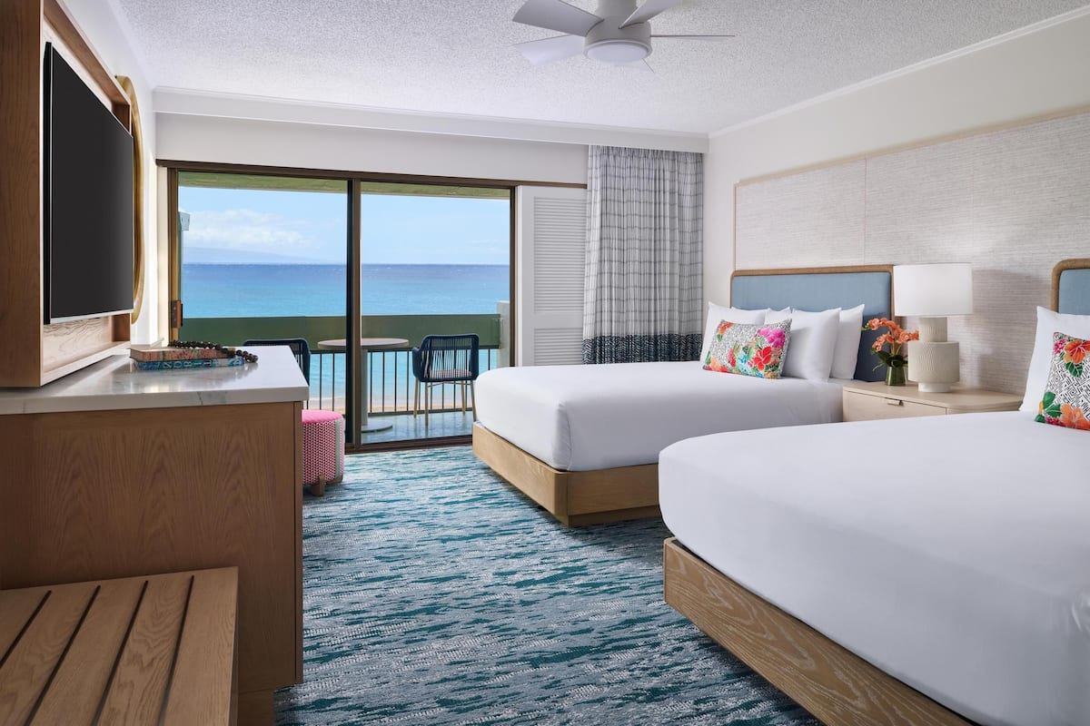 Quarto de hotel com duas camas, televisão, mesa de trabalho, cadeira e varanda vista para o mar.