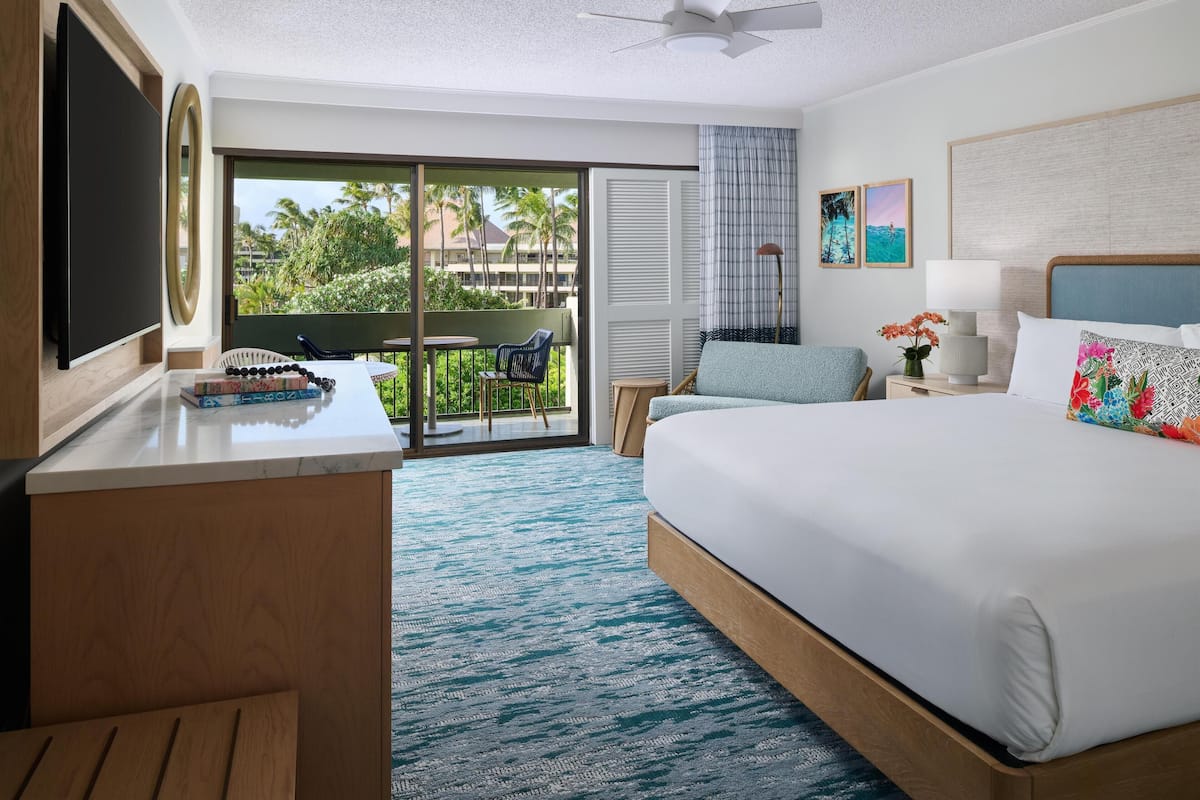 Quarto de hotel com cama, escrivaninha, televisão, varanda e vista para palmeiras, além de janela com cortinas.