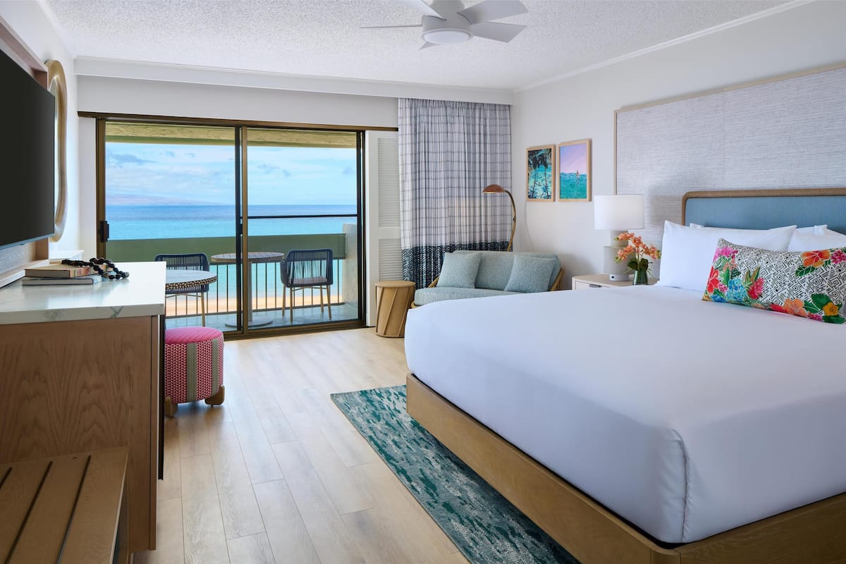 Quarto de hotel com uma cama grande, uma mesa de escritório, uma televisão, varanda com vista para o mar e porta de vidro deslizante.