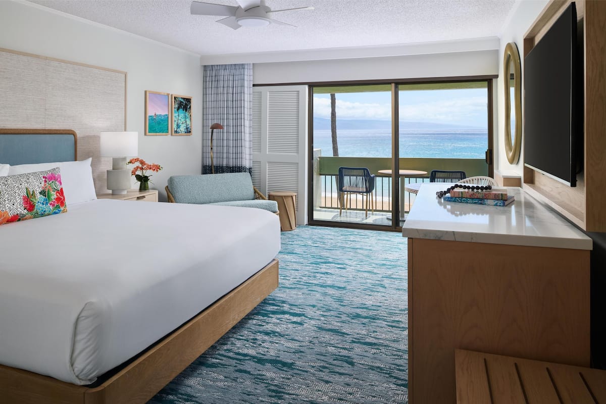 Quarto de hotel com uma cama grande, uma escrivaninha, uma cadeira, uma TV e uma varanda com vista para o oceano.
