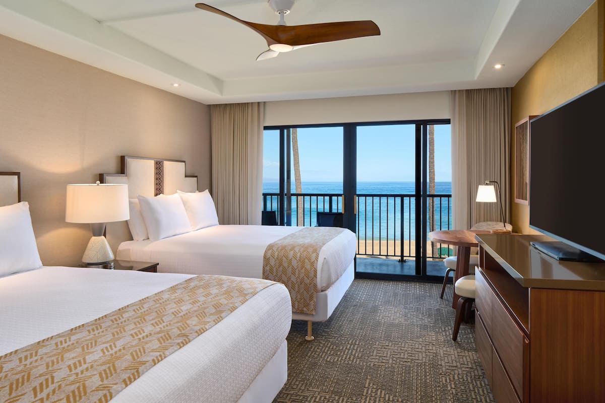 Quarto de hotel com duas camas, uma escrivaninha, televisão, ventoinha no teto e varanda com vista para o mar.