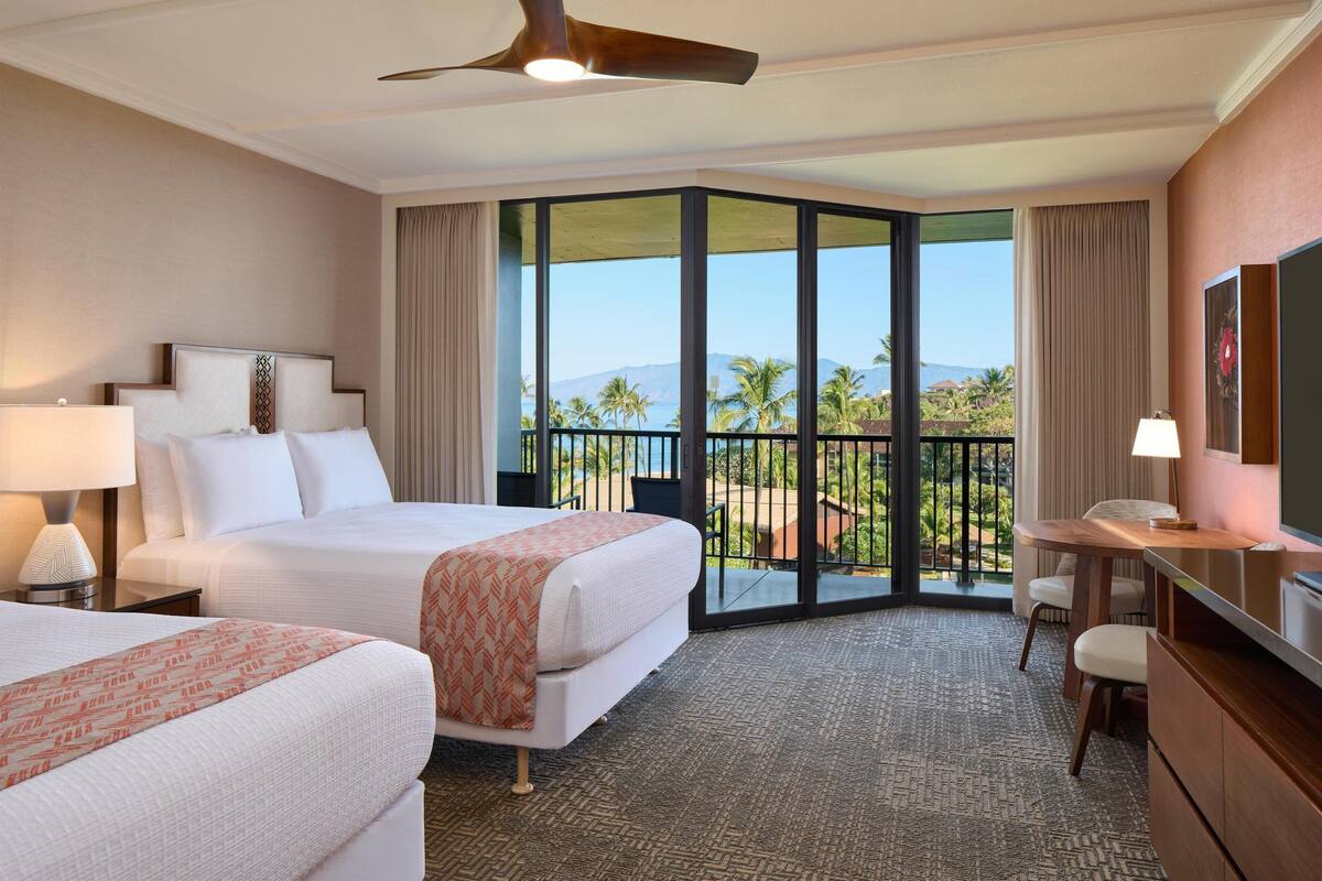 Quarto de hotel com duas camas, uma escrivaninha, uma cadeira, televisão e varanda com vista para palmeiras e montanhas.