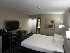 Quarto de hotel com uma cama, uma escrivaninha com televisão, uma lâmpada e um quadro na parede.