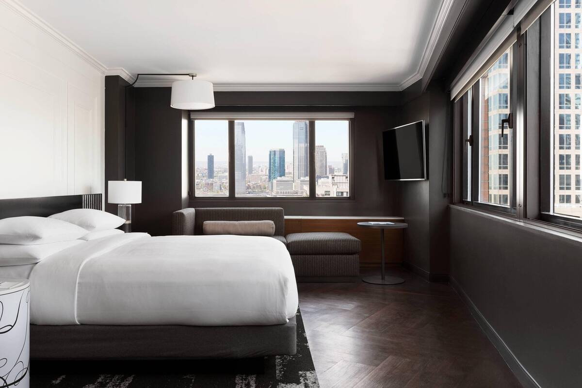 Quarto de hotel moderno com uma cama grande, área de estar, televisão e vista para a cidade.