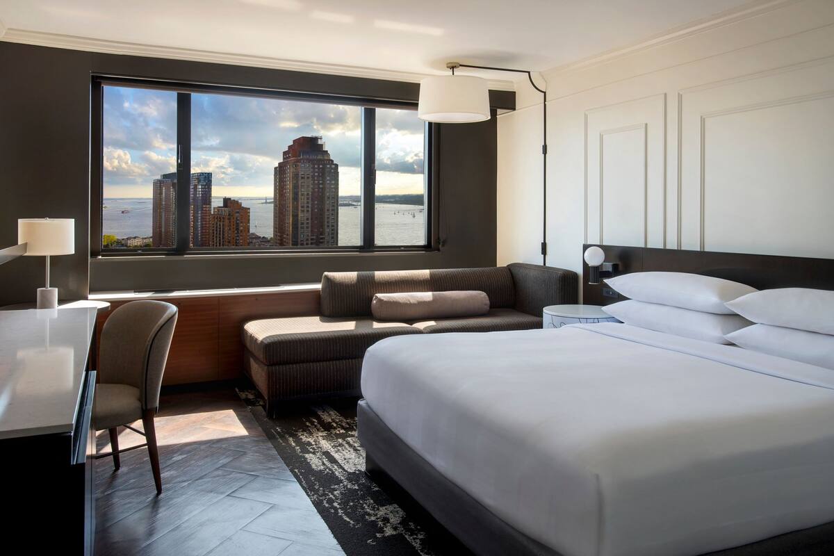 Quarto de hotel com uma janela grande, uma cama, um sofá, uma escrivaninha e uma cadeira.