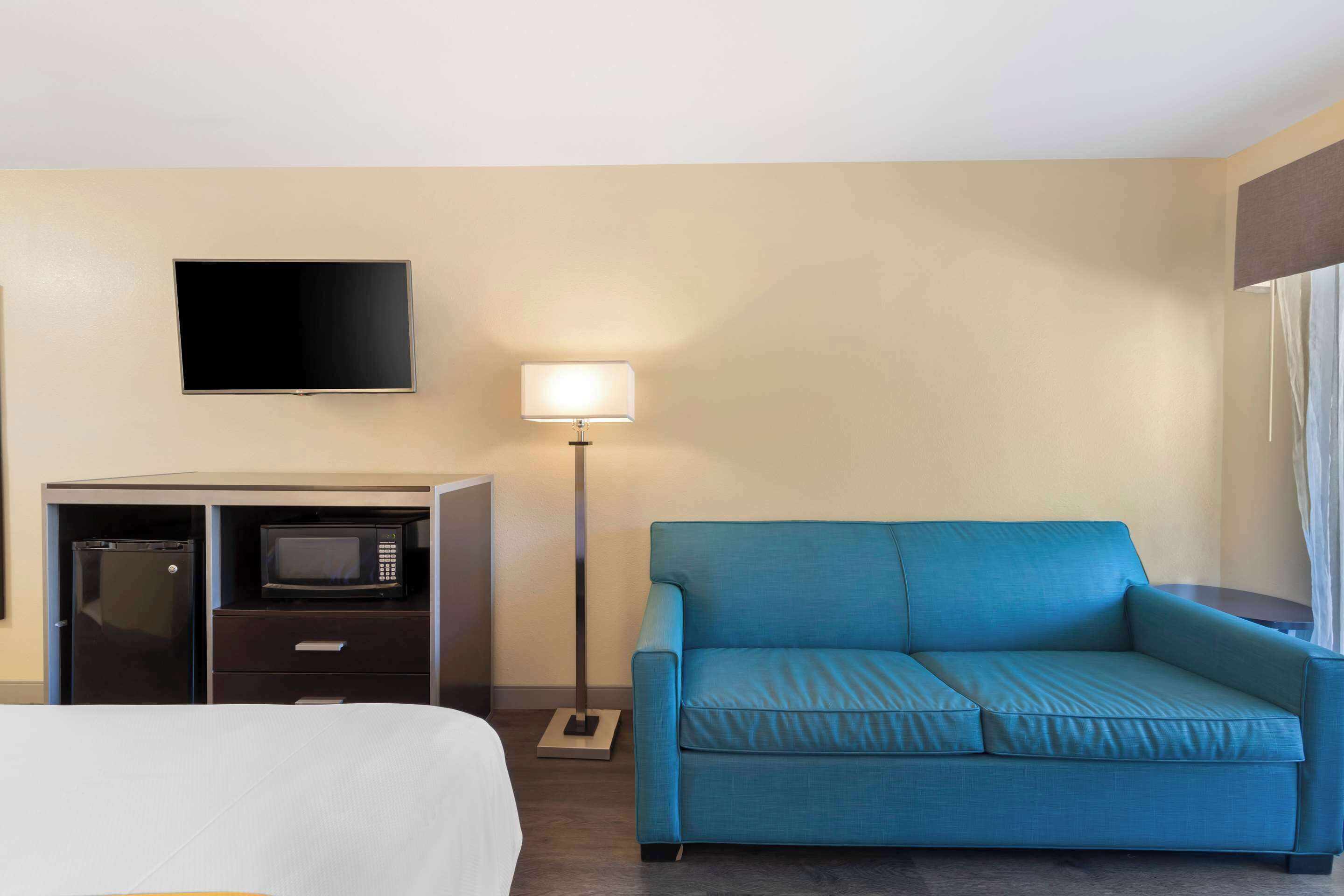 Quarto de hotel com cama, sofá azul, televisão de tela plana, micro-ondas e luminária de chão.