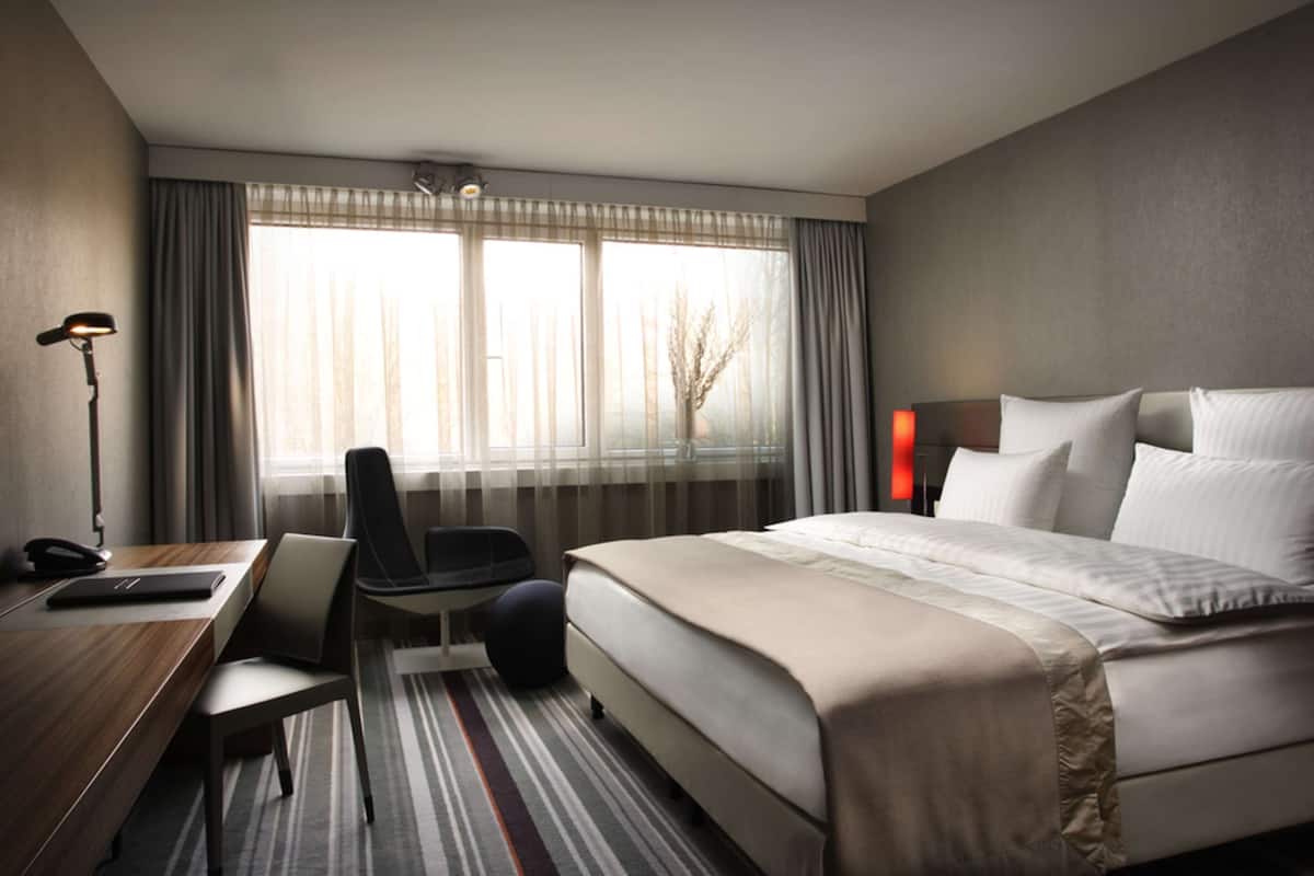 Quarto de hotel com uma cama grande, uma mesa com cadeira, uma luminária e uma janela com cortinas.