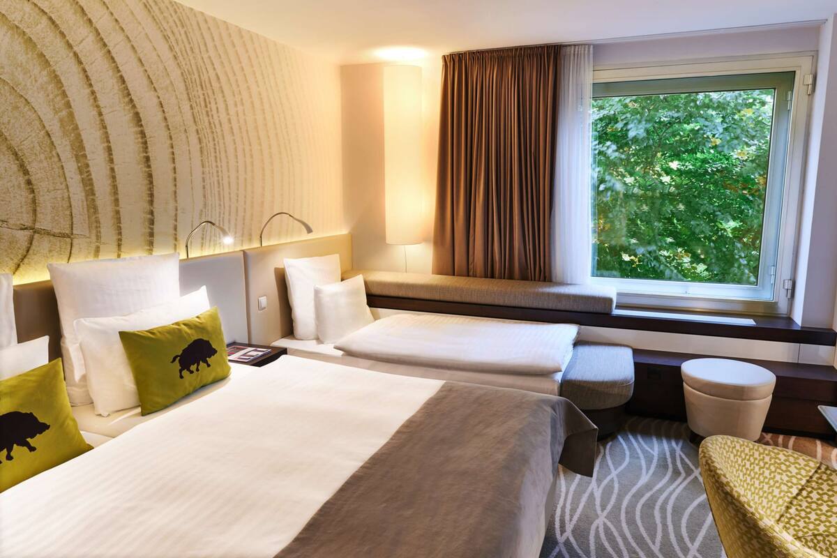 Quarto de hotel com duas camas, uma janela grande com cortinas, cabeceira com design texturizado e um pequeno espaço de estar com um banco e um banquinho.