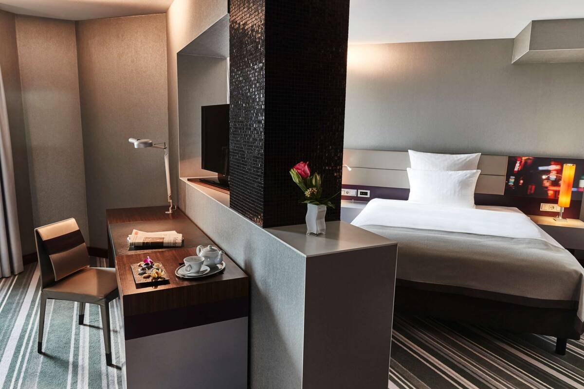 Quarto de hotel moderno com uma cama, uma escrivaninha com cadeira, um vaso com flor e uma televisão.