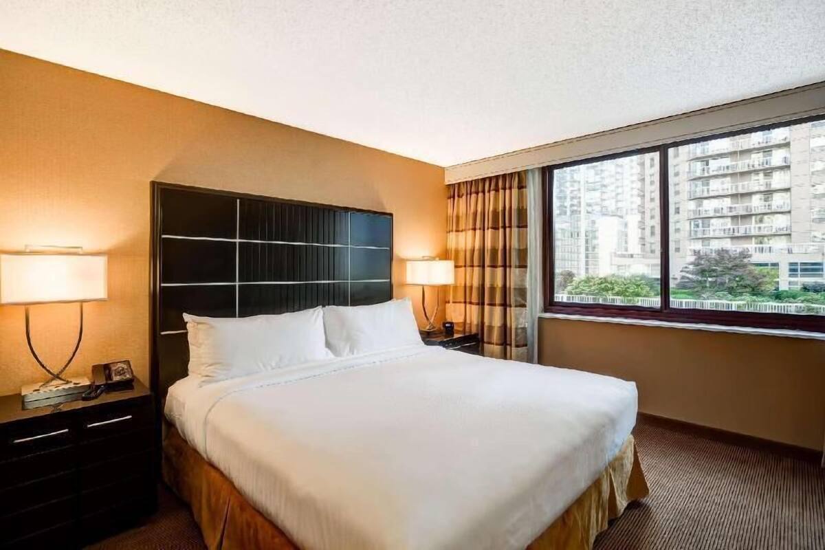 Quarto de hotel com uma cama grande, duas luminárias de cabeceira, uma cabeceira e uma janela com vista para a cidade.