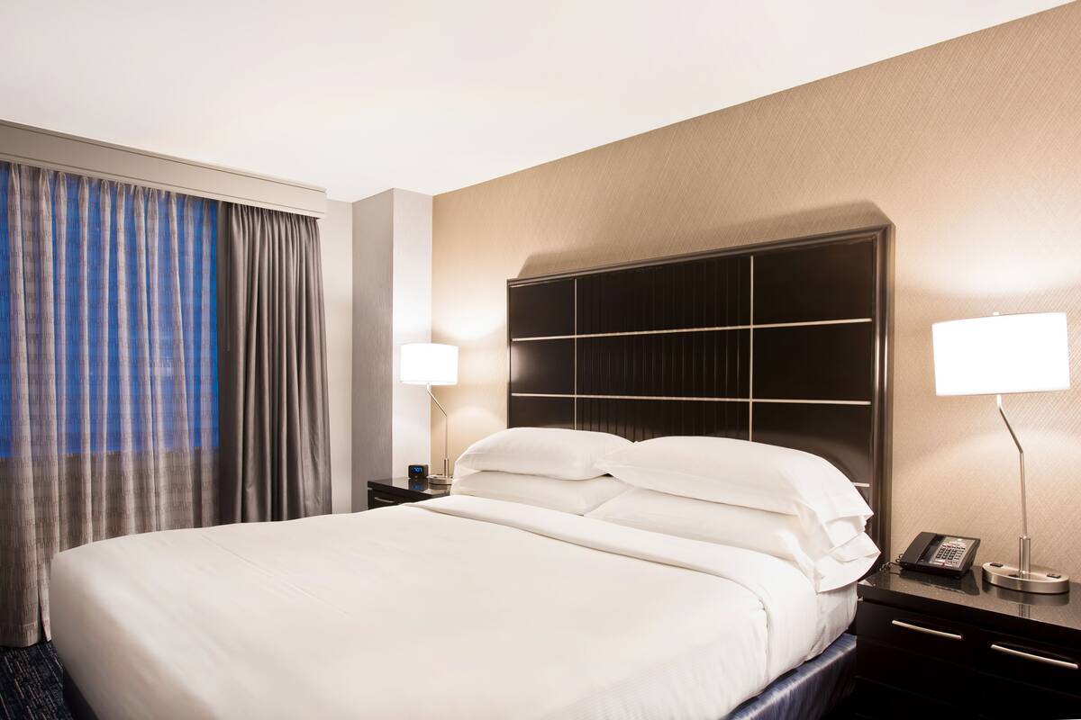 Quarto de hotel com uma cama grande, luminárias de cabeceira, mesa de cabeceira e uma janela com cortinas.