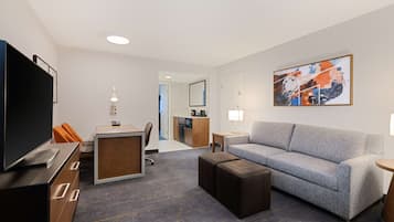 Quarto de hotel com televisão de tela plana, sofá, mesa de centro, escrivaninha com cadeira, cozinha compacta e banheiro visível por uma porta aberta.
