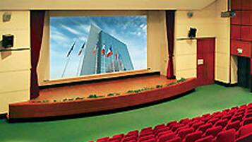 Um teatro com palco, assentos vermelhos e uma tela grande.