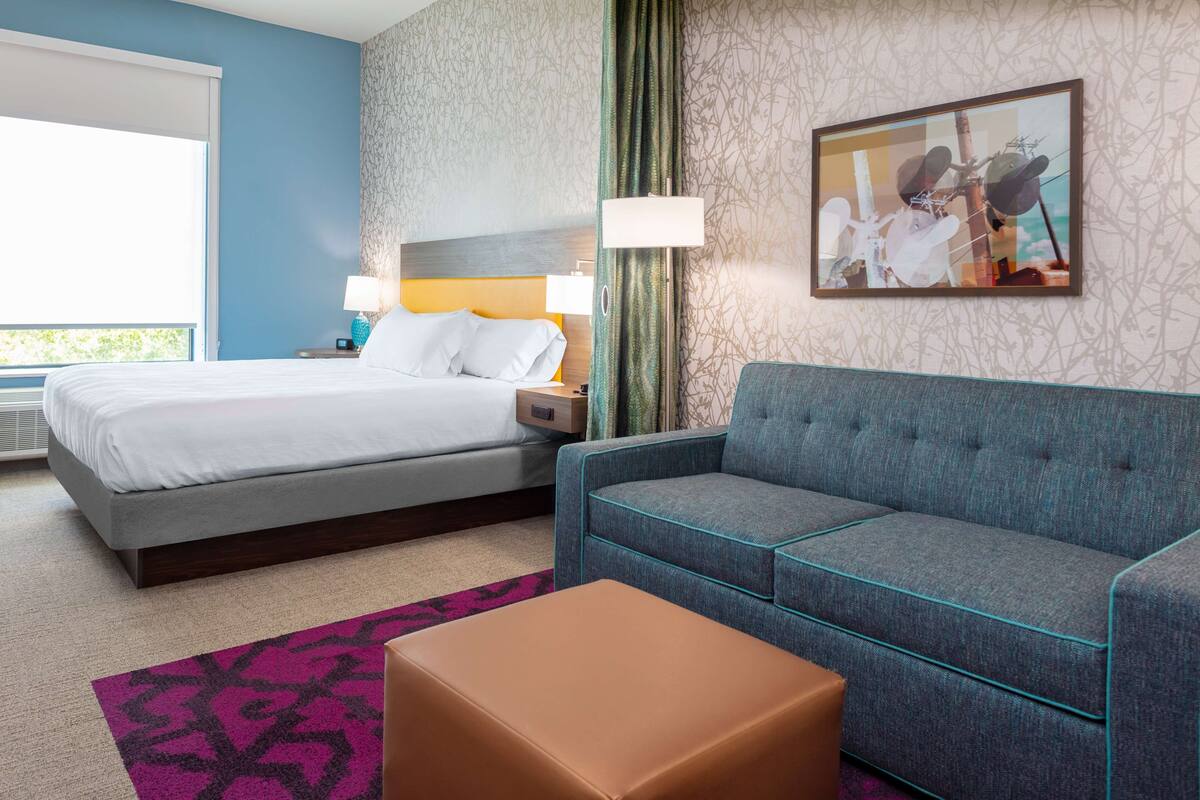 Quarto de hotel com uma cama, um sofá, um pequeno apoio para os pés, uma mesa de cabeceira, um abajur e um quadro na parede.