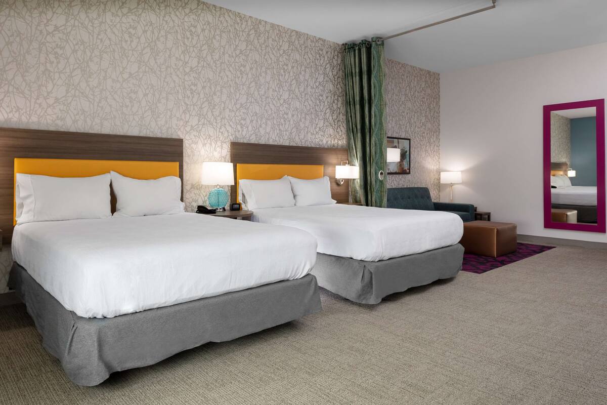 Quarto de hotel com duas camas, um espelho, uma mesinha de cabeceira e cortina verde.