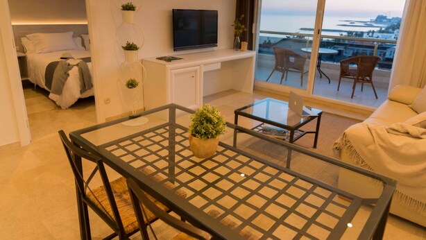 Uma sala de estar moderna com uma mesa de vidro, um sofá, uma TV em um móvel branco e uma varanda com vista para o mar.