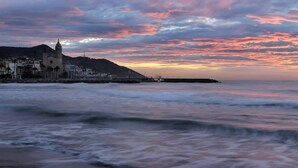 Cena costeira com uma igreja e edifícios ao longo da margem, ondas a aproximar-se da praia, e um céu de pôr do sol.