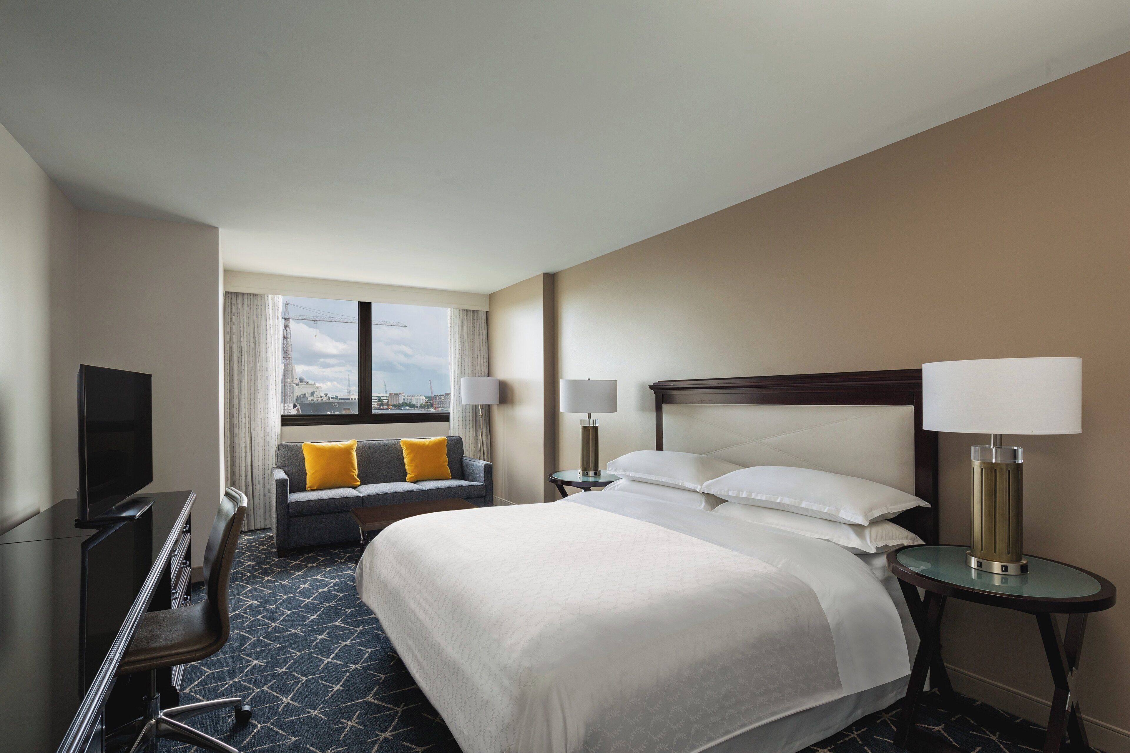 Quarto de hotel com cama, escrivaninha com televisão, sofá com almofadas amarelas, duas luminárias de cabeceira e janela com vista para a cidade.