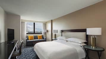 Quarto de hotel com cama, escrivaninha com televisão, sofá com almofadas amarelas, duas luminárias de cabeceira e janela com vista para a cidade.