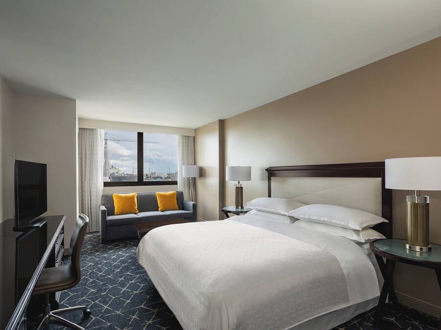Quarto de hotel com cama, escrivaninha com televisão, sofá com almofadas amarelas, duas luminárias de cabeceira e janela com vista para a cidade.