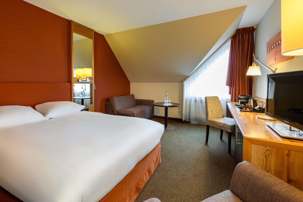 Quarto de hotel com uma cama grande, uma escrivaninha com televisão, uma cadeira e uma mesinha com garrafa e copos.