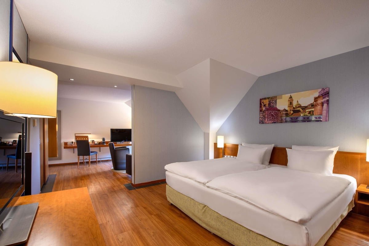 Quarto de hotel com uma cama grande, uma escrivaninha com cadeira, uma televisão e um quadro na parede.