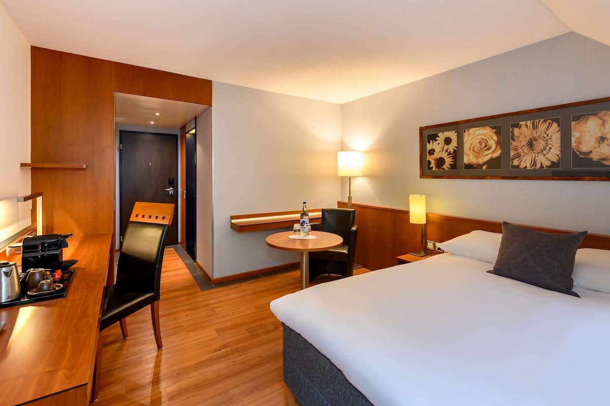 Quarto de hotel com uma cama, uma escrivaninha com cadeira, uma mesinha com cadeira e uma luminária na parede.