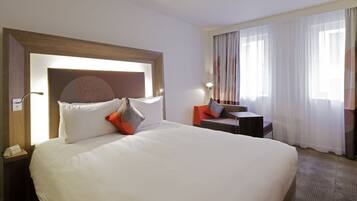 Quarto de hotel com uma cama grande, uma mesa e vista para o exterior através de cortinas leves.