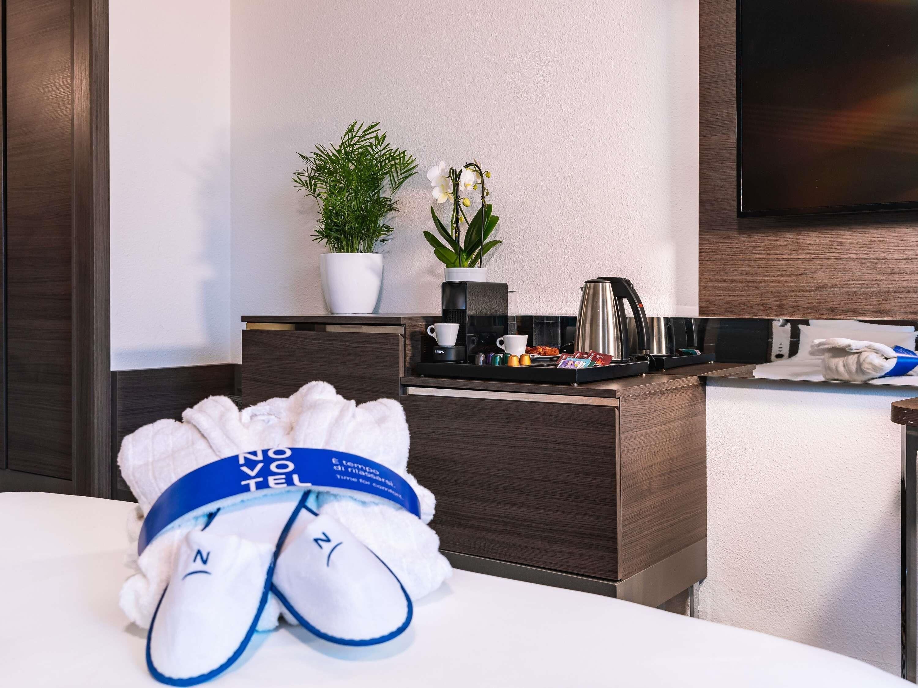 Quarto de hotel com cama, mesa de cabeceira com toalha e chinelos, cafeteira e uma planta.