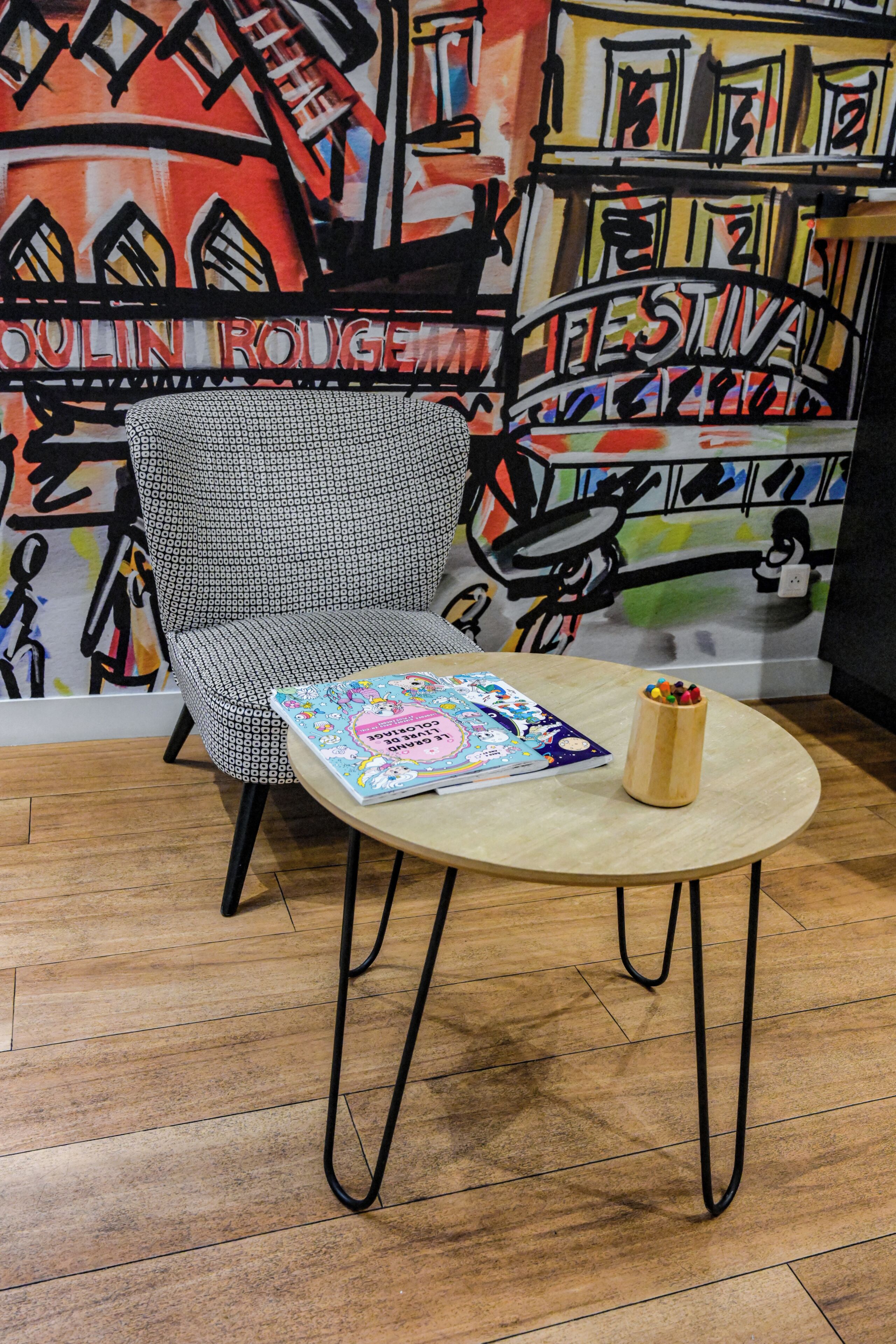 Um quarto com uma cadeira estampada, uma pequena mesa redonda com um livro e uma xícara, e um mural colorido na parede.