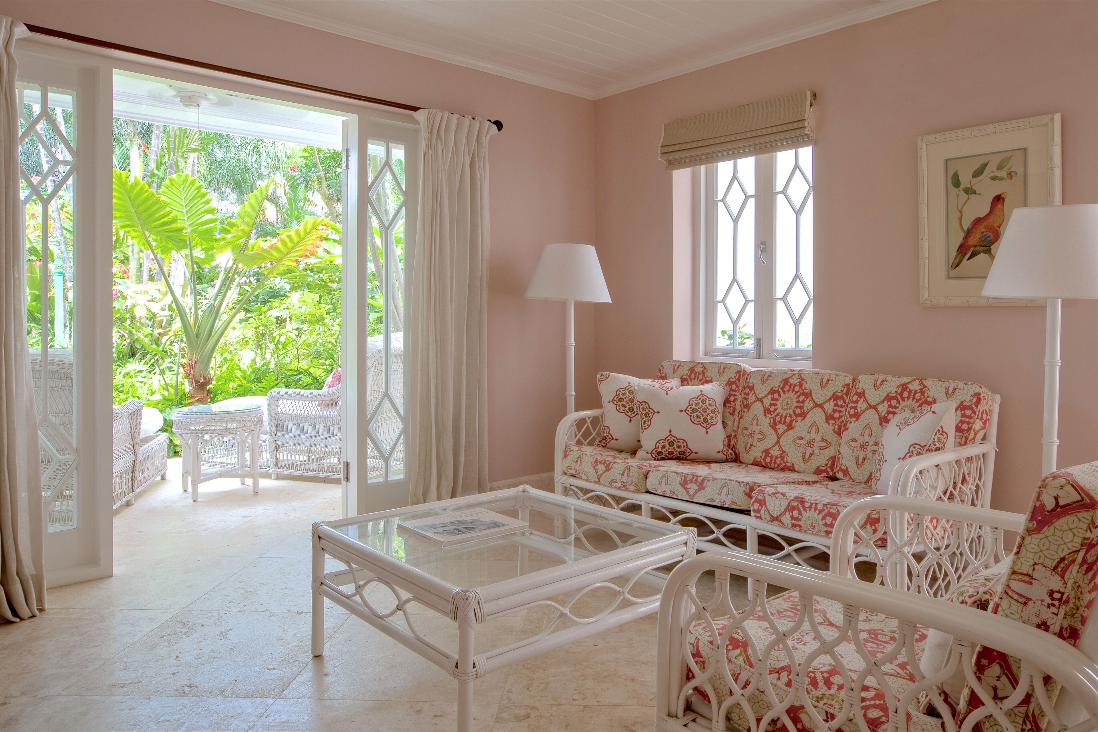 Quarto com sofá branco, mesa de centro de vidro e cadeiras de vime. Janela com vista para o verde lá fora.