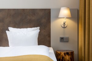 Cama bem arrumada com lençóis brancos e cabeceira de madeira, luminária fixada na parede e mesinha de cabeceira de madeira.