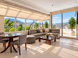 Uma sala de estar moderna com sofá, mesa de centro e cadeiras, janelas amplas com vista para a cidade e um vaso de planta.