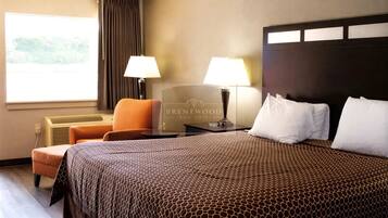 Quarto de hotel com uma cama grande, duas luminárias de cabeceira, uma cadeira, uma mesa pequena e uma janela com cortinas.