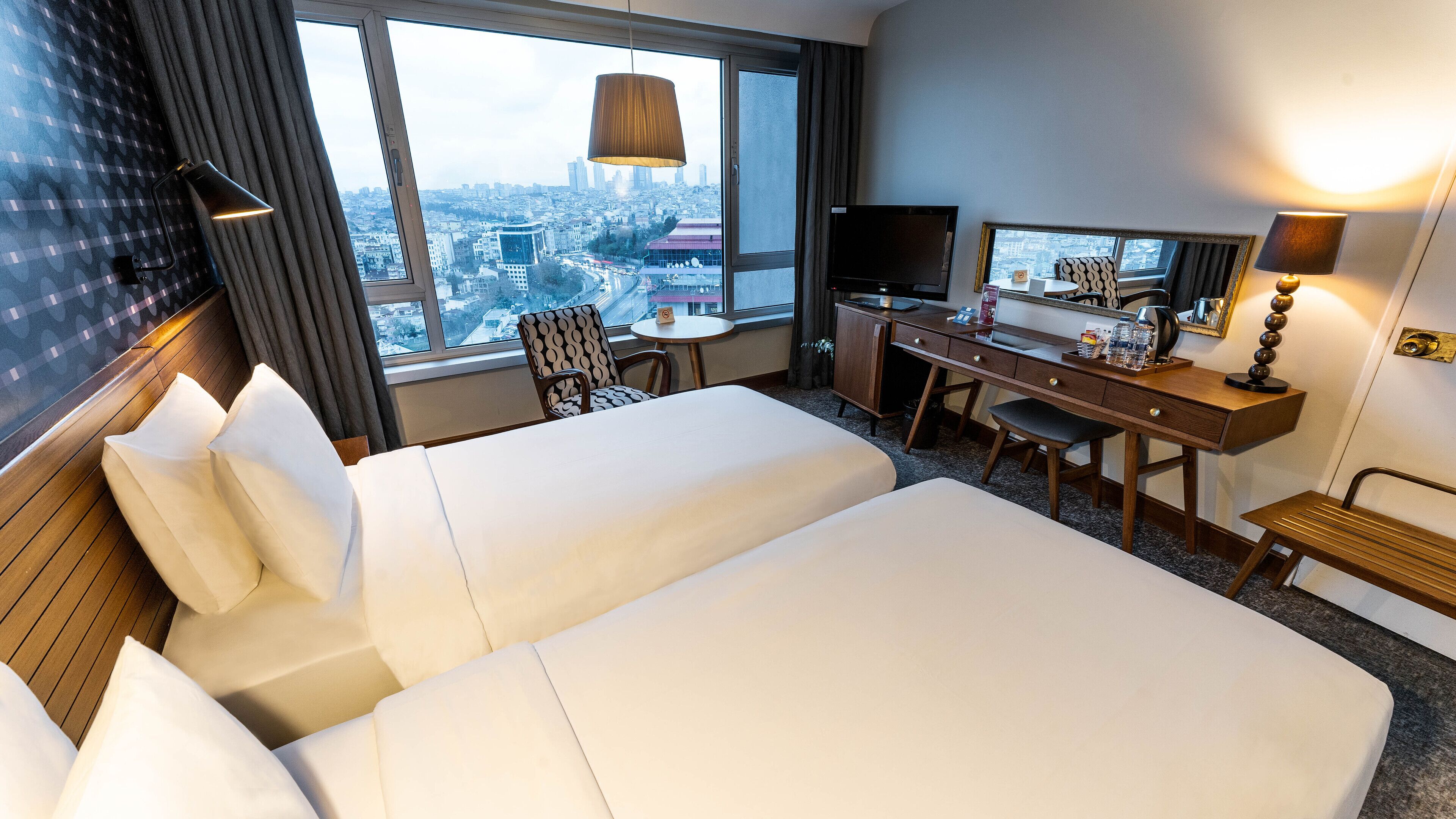 Quarto de hotel com uma cama grande, uma escrivaninha com um computador, uma cadeira e vista para a cidade através de uma janela ampla.