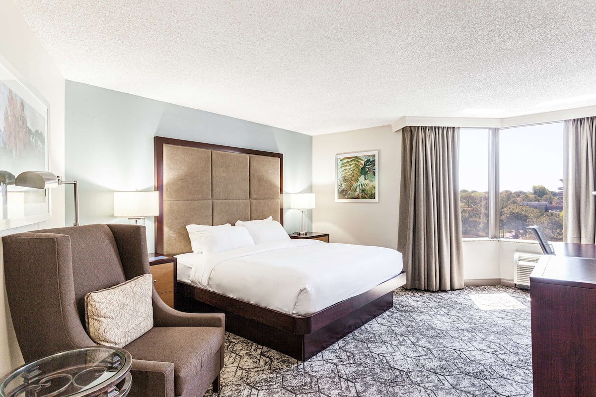 Quarto de hotel com cama, poltrona, escrivaninha e uma janela grande com cortinas.